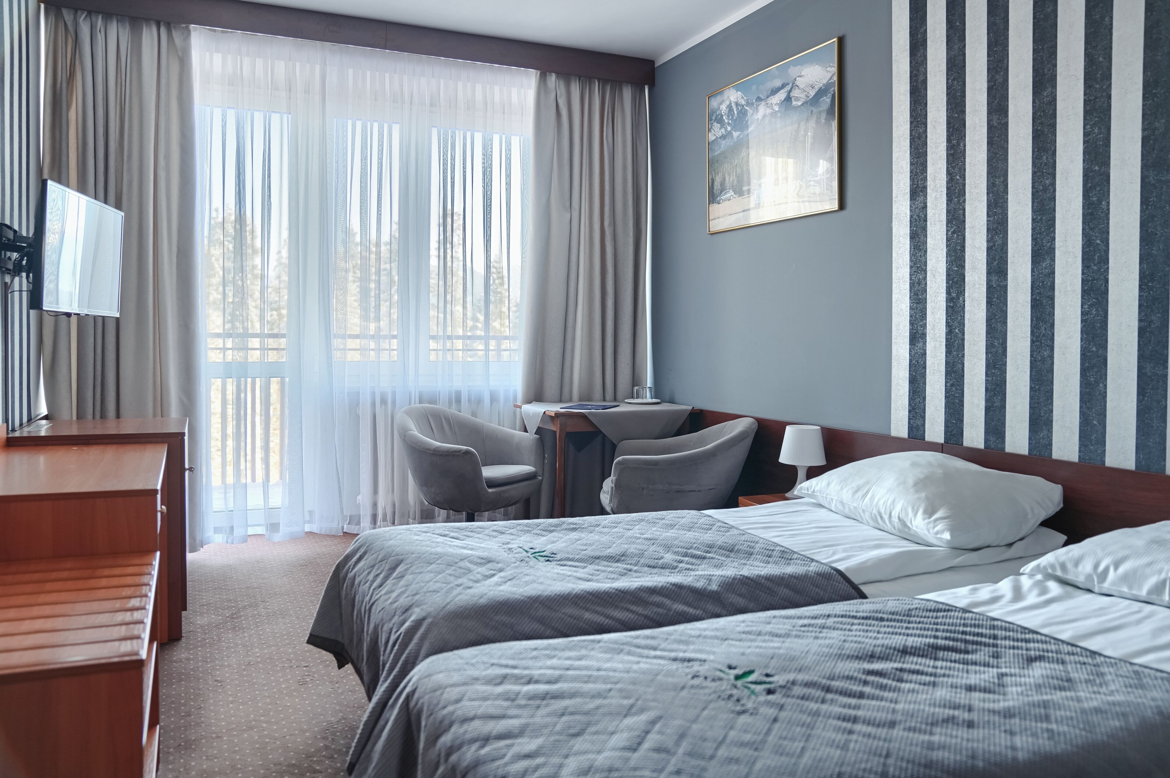 Quarto de hotel com duas camas, uma escrivaninha, uma cadeira e uma janela grande com cortinas.