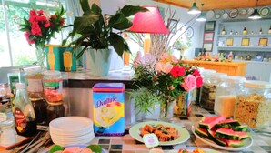 Uma mesa de buffet com diversos tipos de comida, incluindo pratos com carne fatiada, camarões e fatias de melancia. Há também potes de condimentos e uma caixa de Ovaltine.