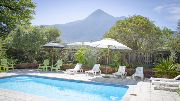 Piscina com espreguiçadeiras e guarda-sóis, cercada por um muro de madeira e vegetação, com uma montanha ao fundo.