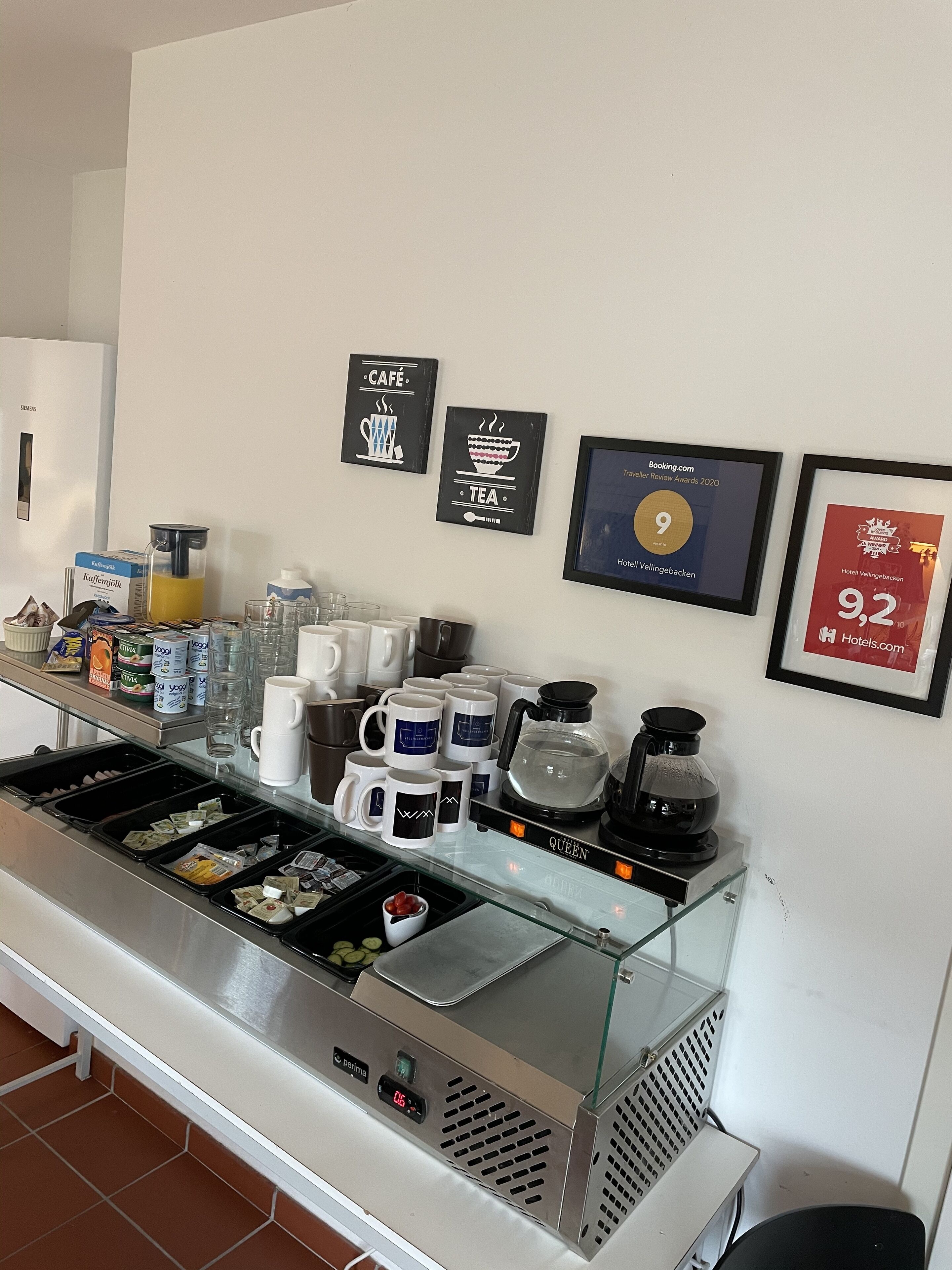 Uma mesa de café da manhã com opções de café e chá, uma geladeira e prêmios emoldurados na parede.