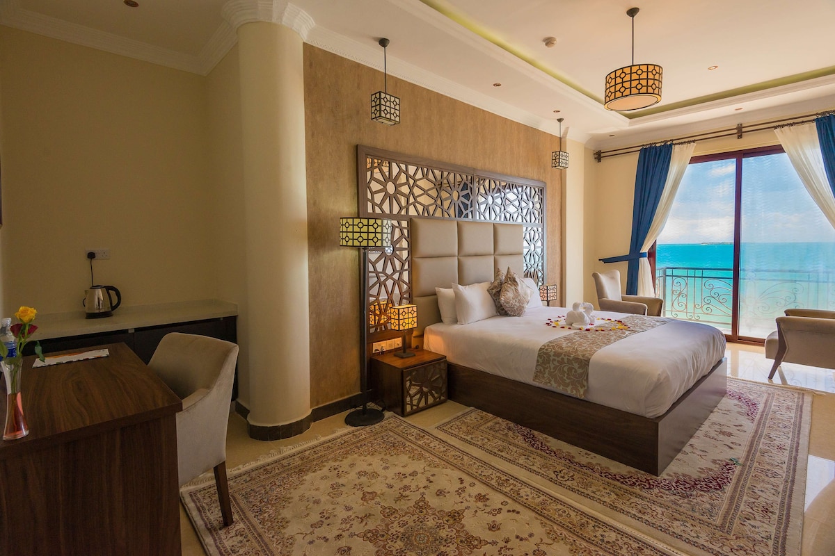 Quarto de hotel com cama grande, escrivaninha, cadeira, mesa de cabeceira e vista para o mar.