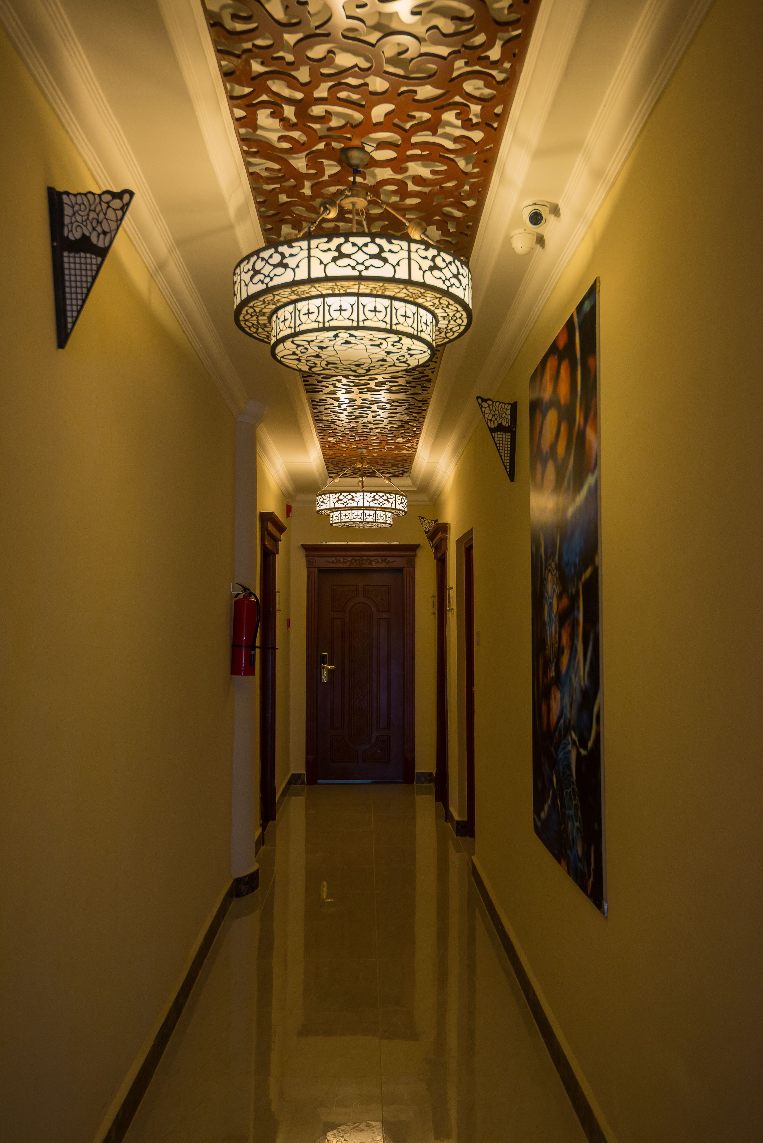 Um corredor com teto decorativo, um lustre, um extintor de incêndio e um quadro na parede.