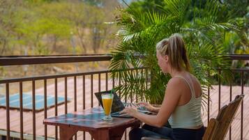 Uma pessoa usando um laptop em uma mesa de madeira, com uma bebida sobre ela, ao ar livre, com grades e vegetação ao fundo.