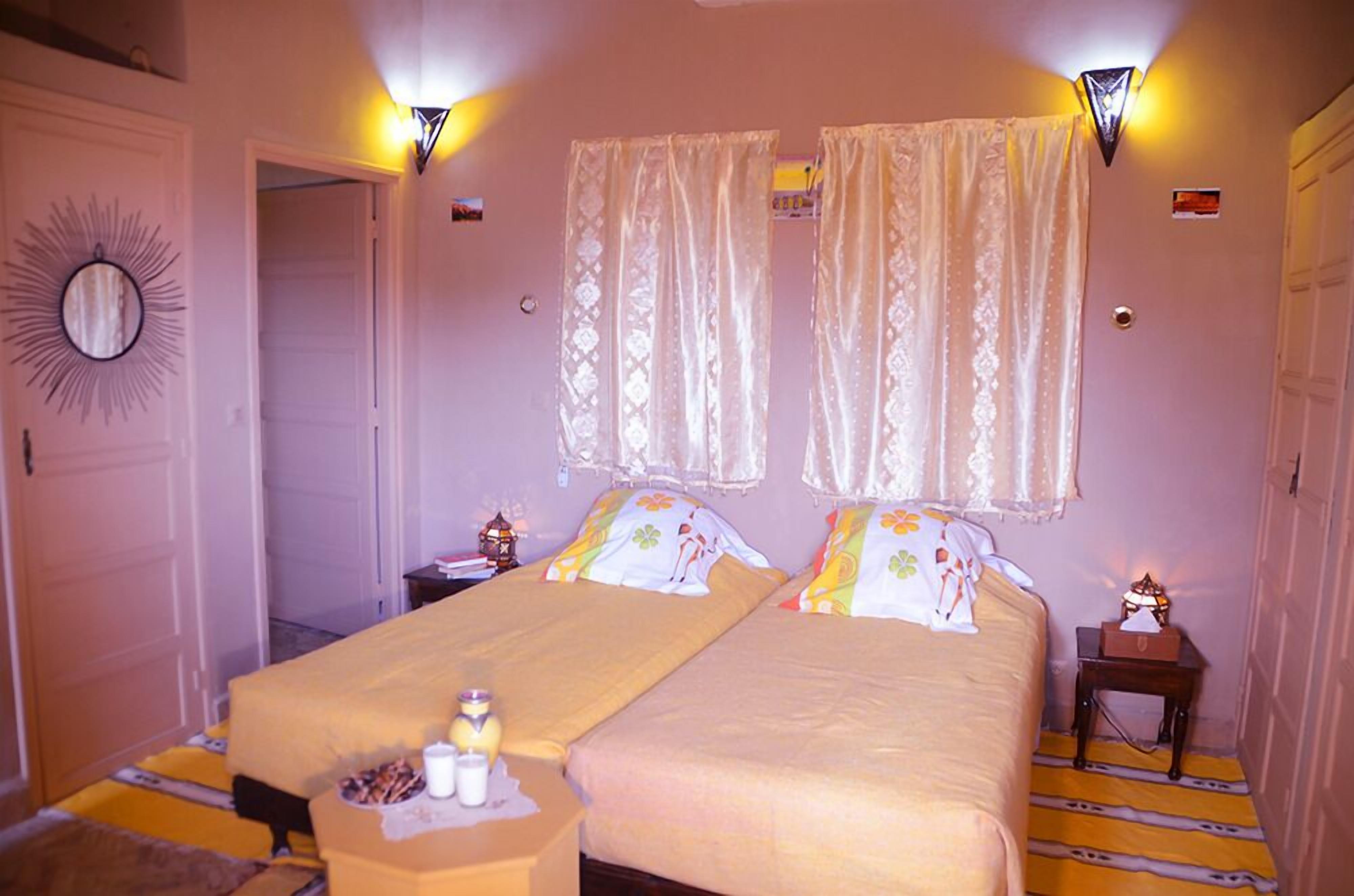 Quarto com duas camas, uma mesinha de cabeceira, um espelho e uma janela com cortinas.