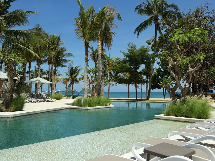 Área de piscina com água cristalina, cercada por palmeiras e espreguiçadeiras, com vista para uma praia e oceano.