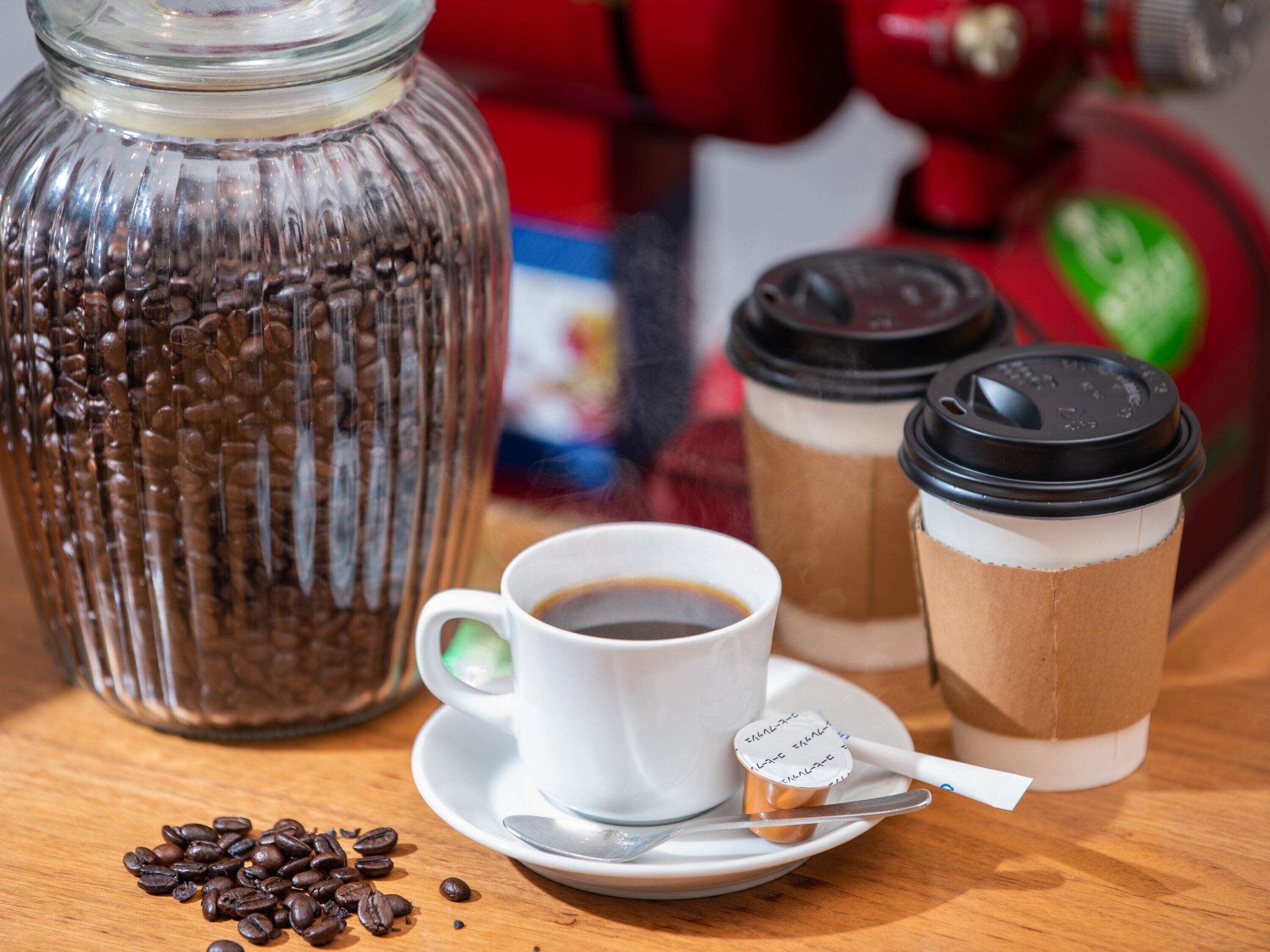 Uma composição com café, uma xícara, um pote de grãos de café e duas xícaras descartáveis sobre uma mesa de madeira.