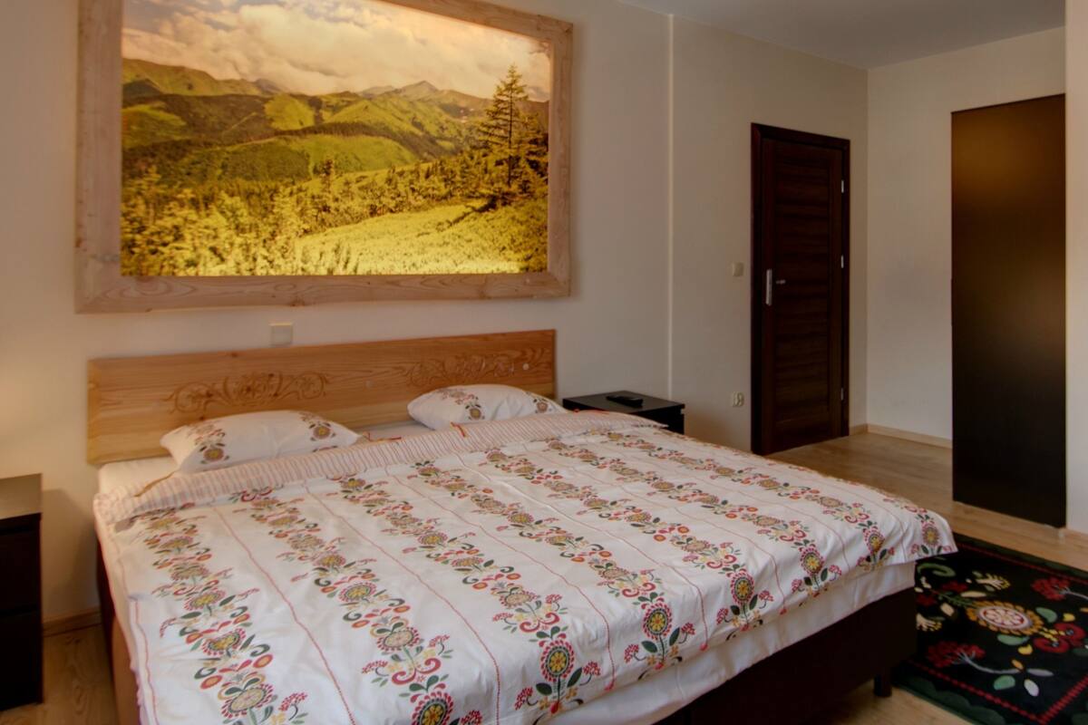 Quarto com uma cama grande, mesa de cabeceira, cabeceira de madeira, pintura na parede, porta e tapete.