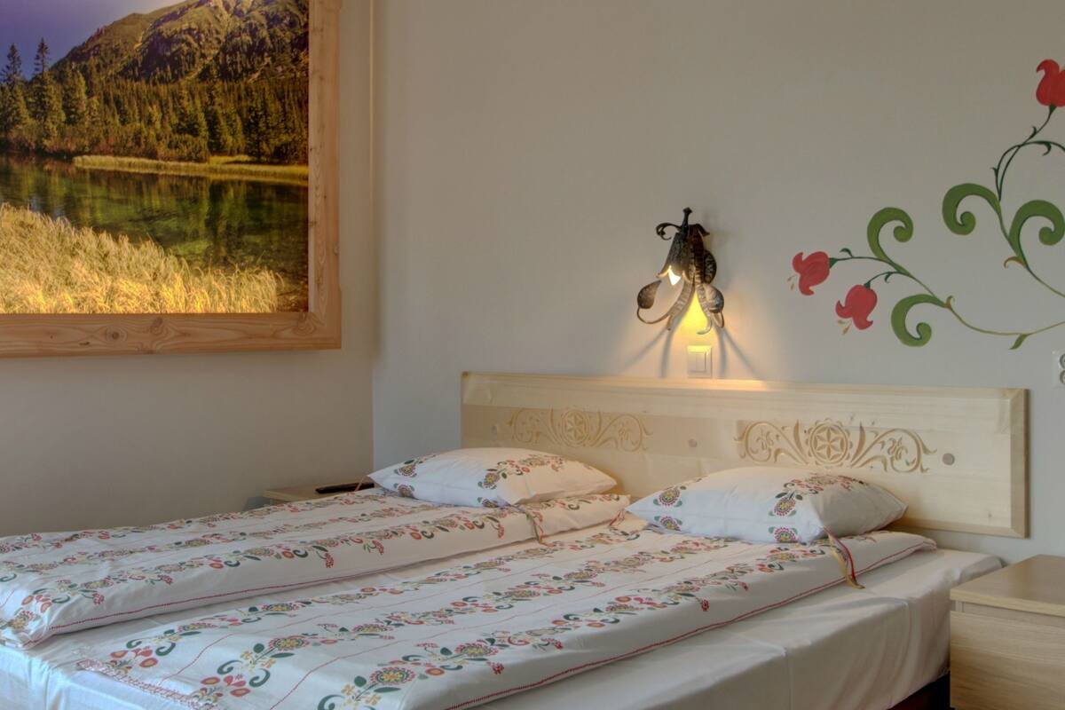 Quarto com cama, mesa de cabeceira, luminária na parede e pintura de uma paisagem de montanhas.