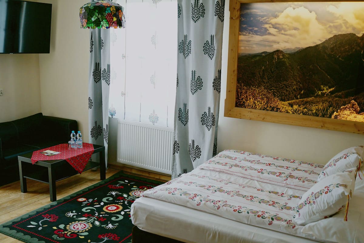 Quarto com cama, mesa com garrafas de água, toalha de mesa vermelha, tapete estampado e pintura de paisagem de montanhas.