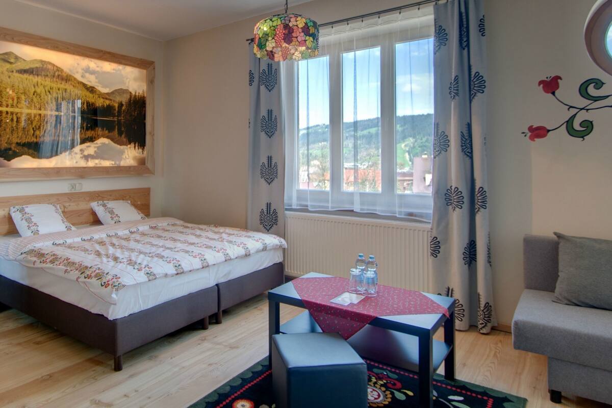 Quarto com cama, mesa com duas garrafas de água, sofá e janela com cortinas.