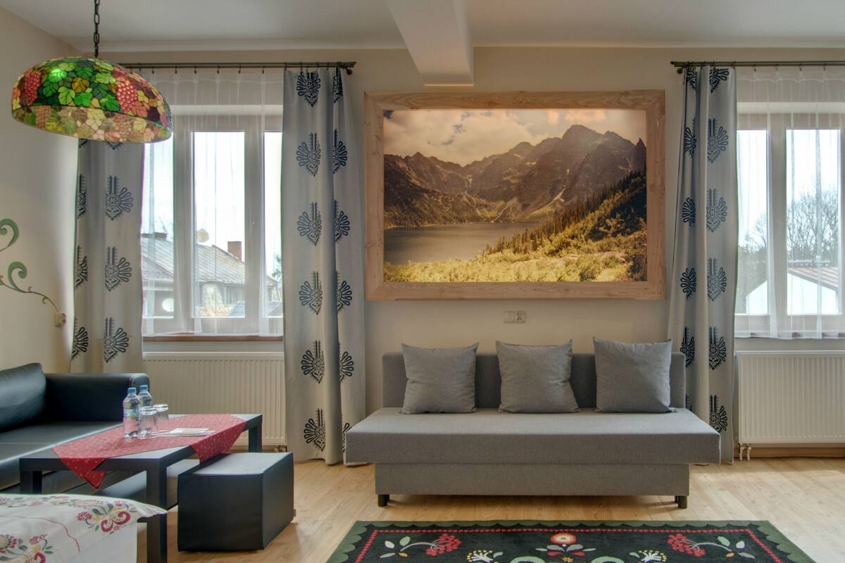 Uma sala de estar moderna com um sofá cinzento, uma mesa vermelha com toalha de mesa florida, um candeeiro pendente e um grande quadro de uma paisagem montanhosa na parede.