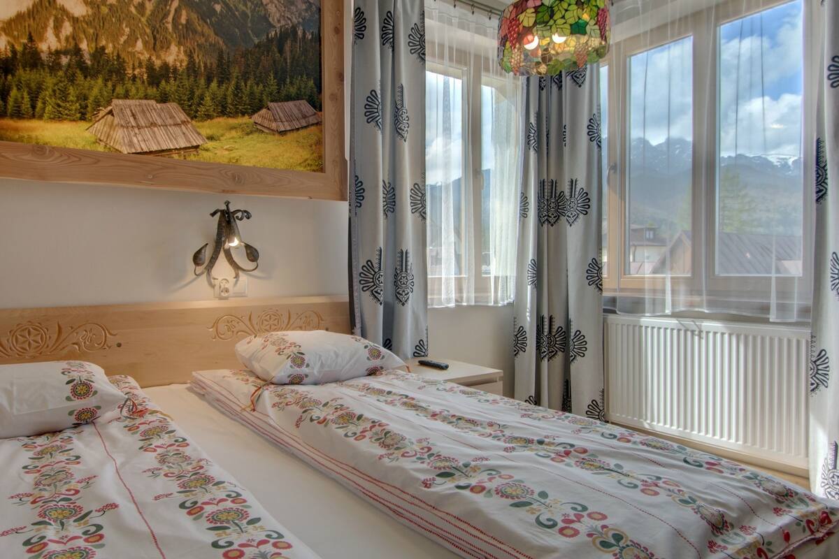 Quarto com cama, mesa de cabeceira, luminária na parede e janela com cortinas.
