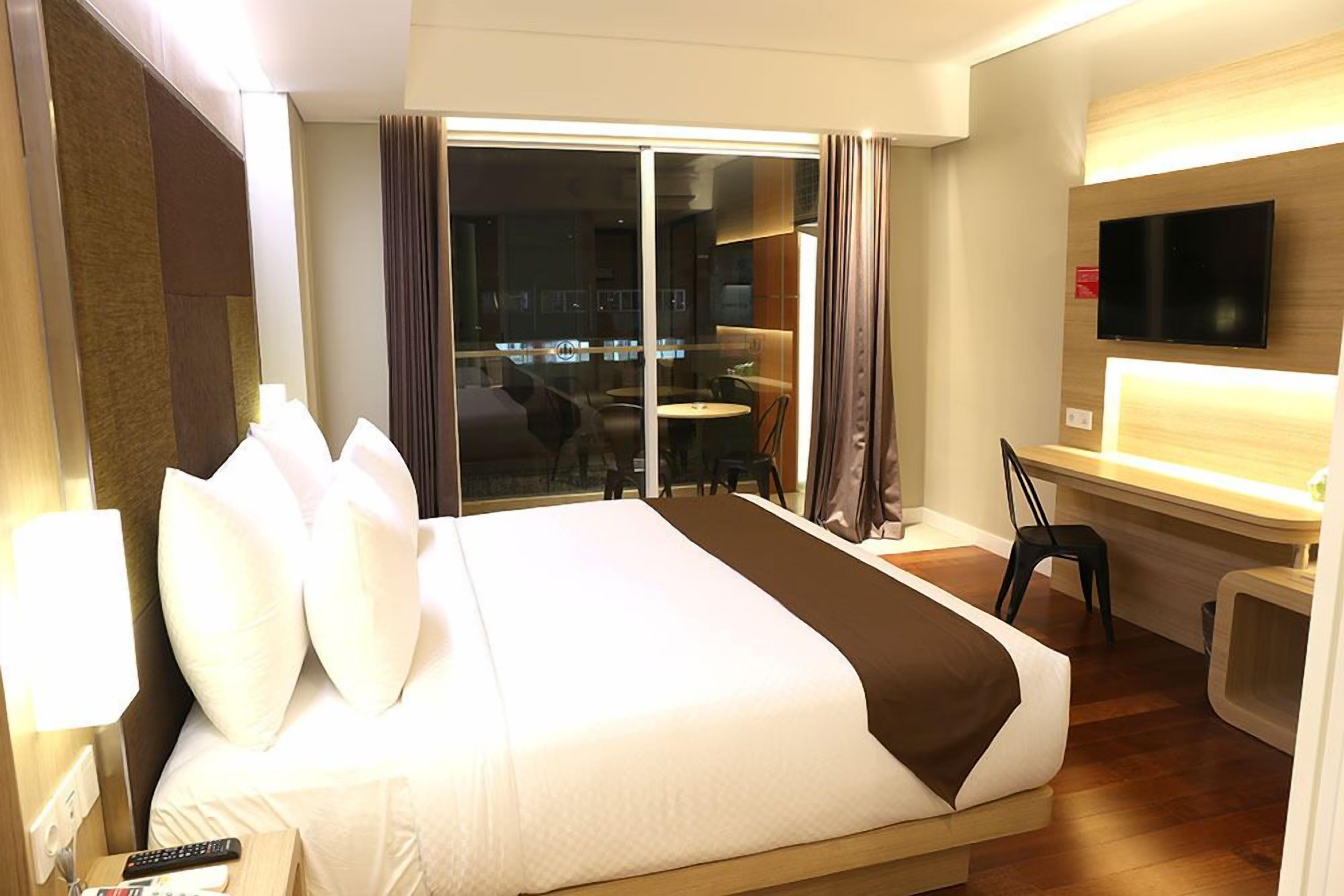 Quarto de hotel com uma cama grande, uma escrivaninha com cadeira, uma televisão na parede e uma porta de correr para uma área de refeições.