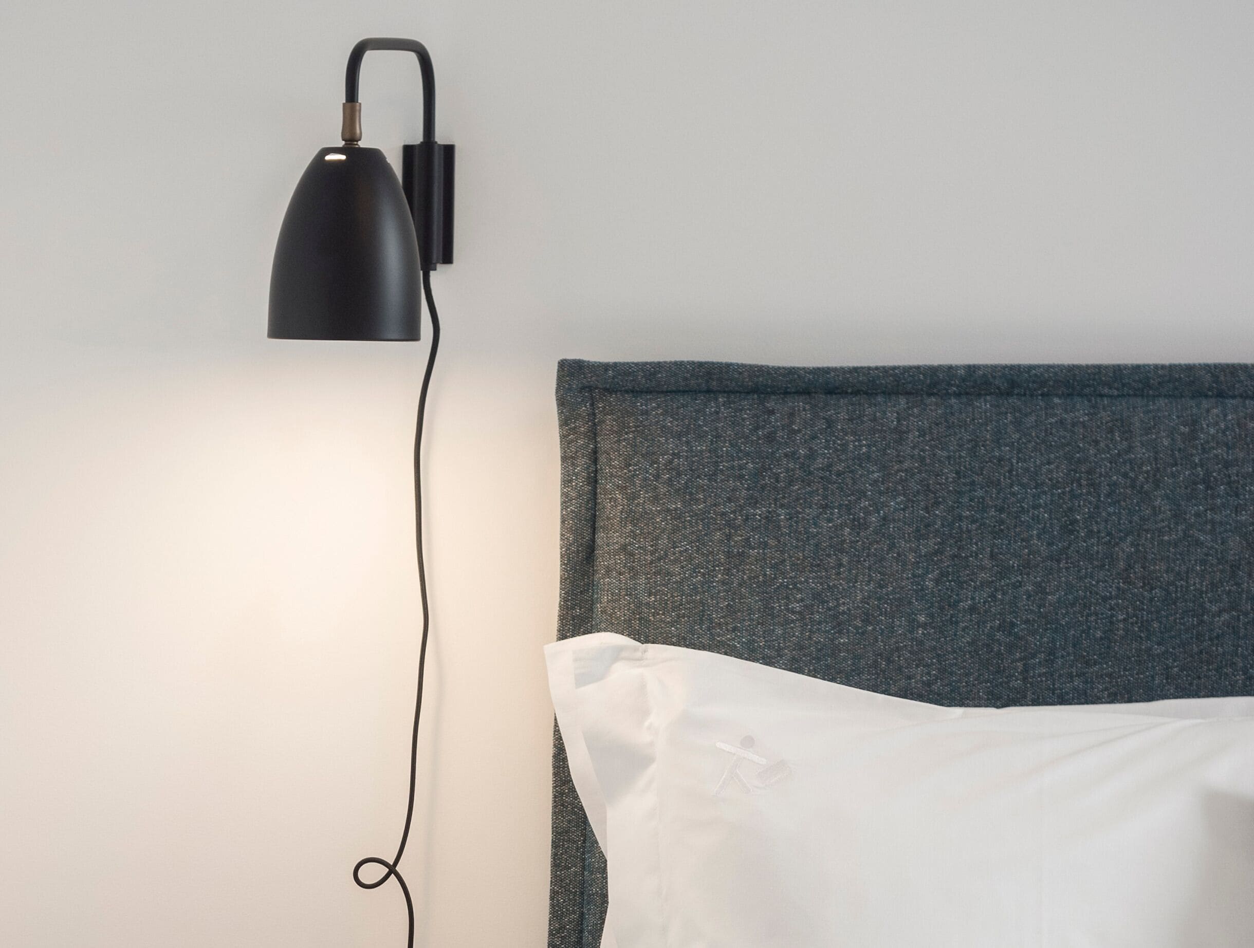 Uma cama com travesseiro branco e cabeceira escura, uma luminária de mesa preta e uma parede lisa.