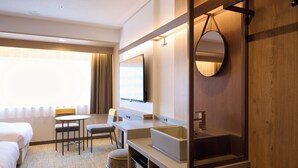 Quarto de hotel com uma cama, uma escrivaninha, uma cadeira, uma mesinha, uma pia, um espelho e um cesto de lixo.