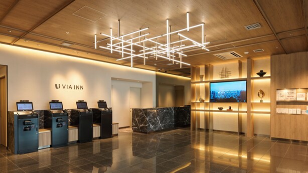 Uma área de recepção moderna, com balcão de mármore, totens digitais de check-in e uma tela grande exibindo o nome do hotel.