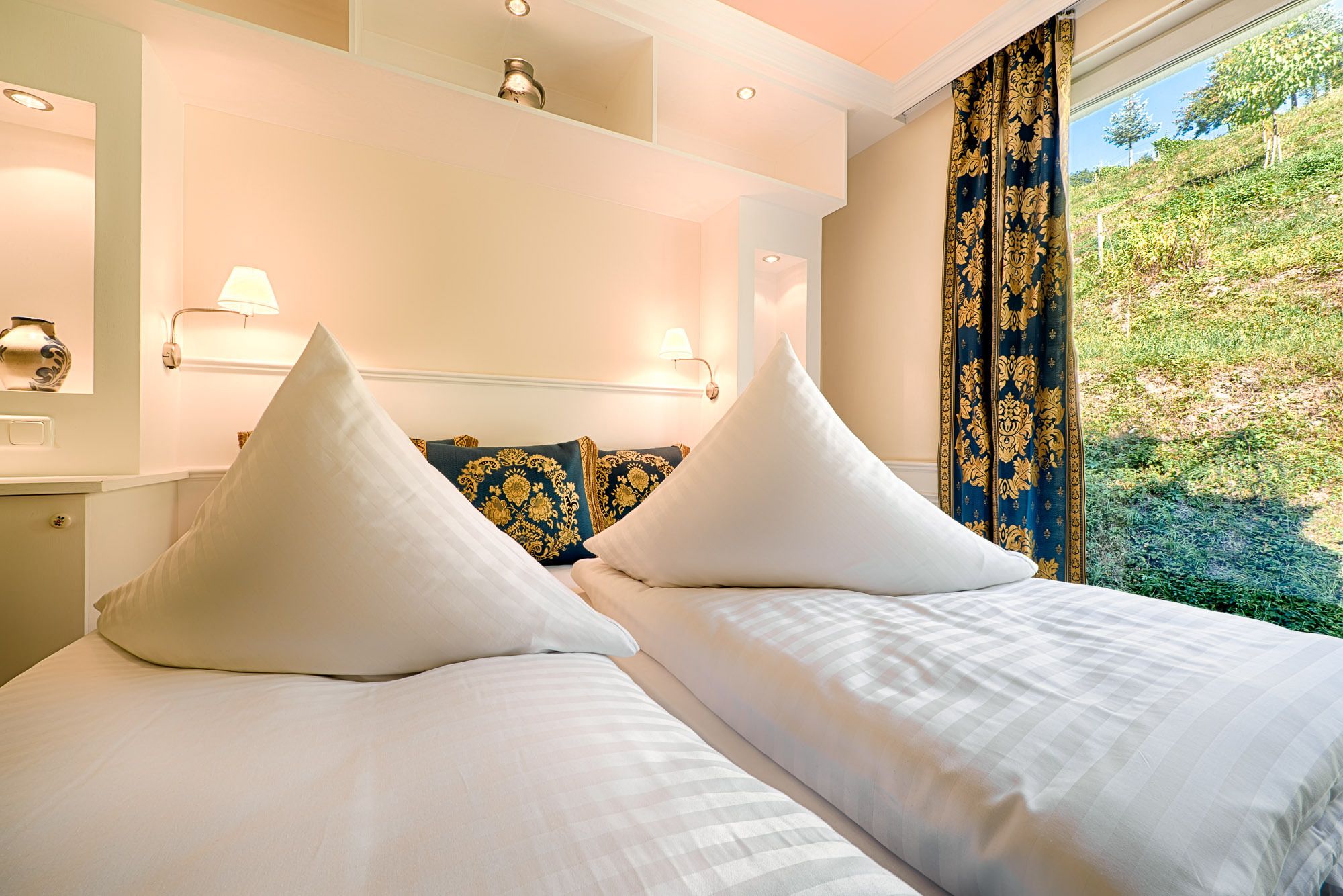Quarto de hotel com duas camas, cada uma com roupa de cama branca e uma almofada decorativa. Há luminárias na parede e vista para o verde lá fora.