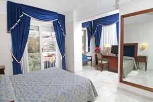 Quarto de hotel com cama, escrivaninha, cadeira, espelho e varanda com cortinas azuis.