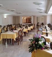 Uma sala de banquetes com várias mesas redondas, cada uma coberta por uma toalha amarela e rodeada por cadeiras de madeira.