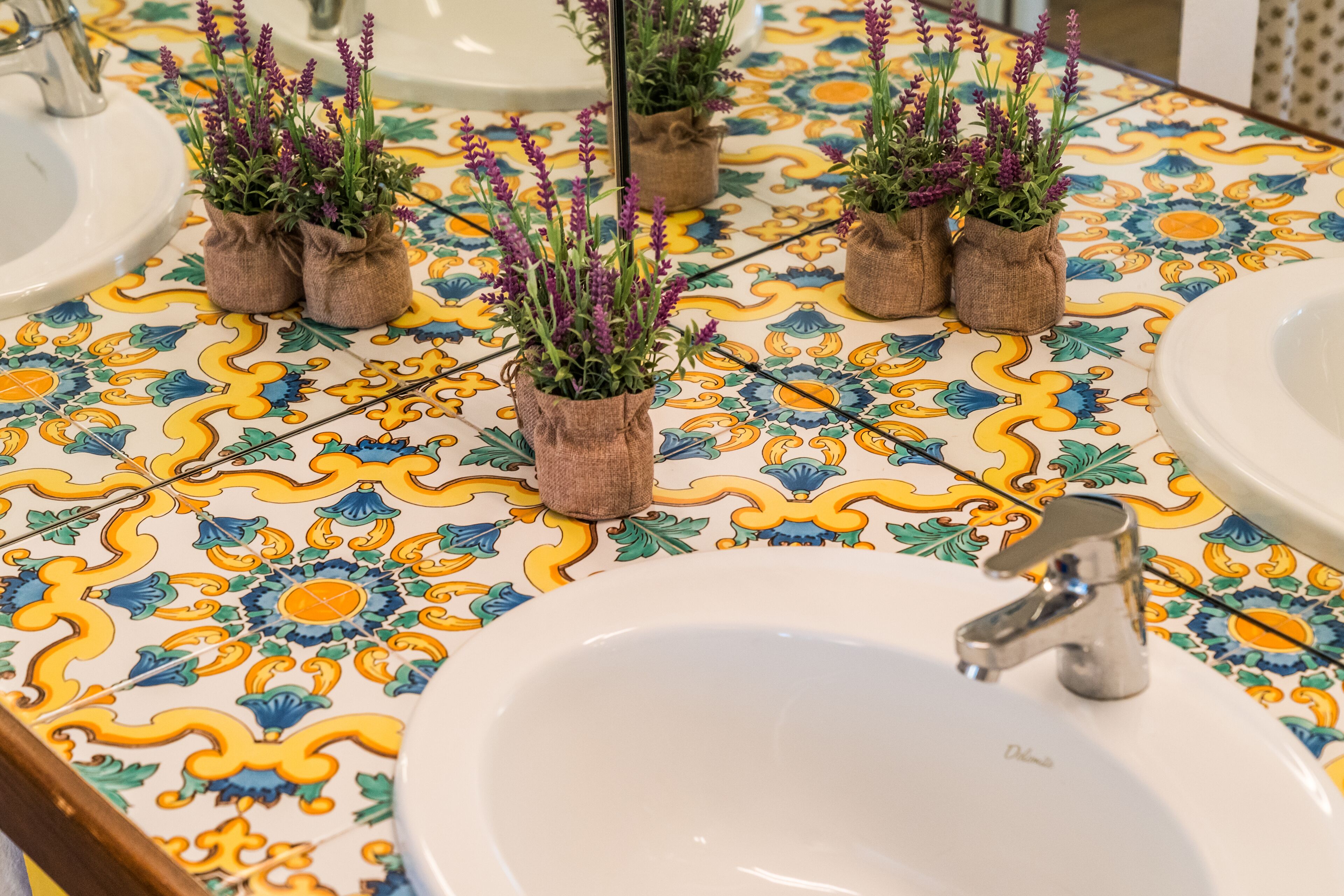Um banheiro com piso ladrilhado, um padrão floral colorido, várias pias e pequenos vasos com flores roxas.