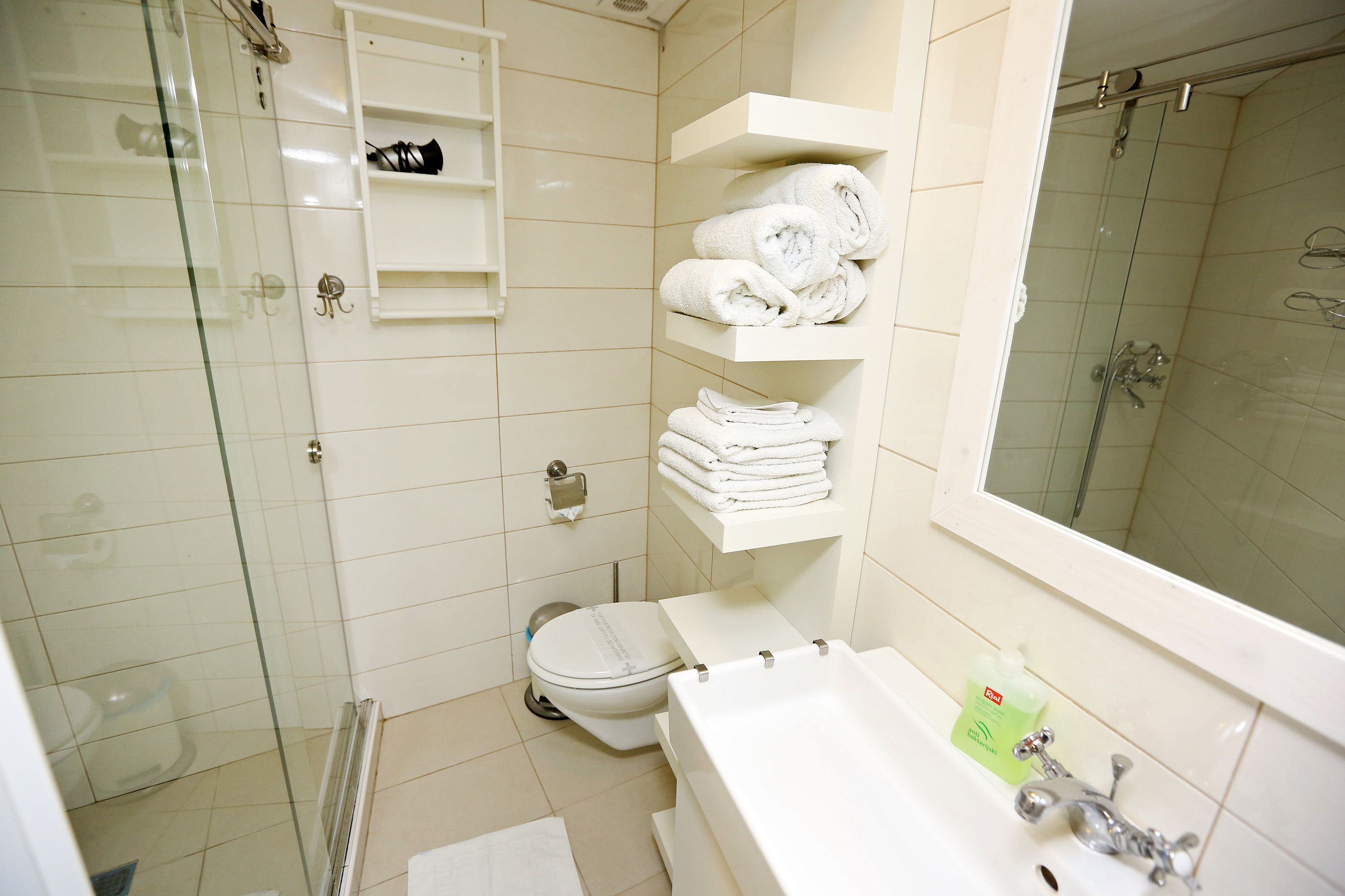 Casa de banho com cabine de duche em vidro, sanita, lavatório com espelho e prateleiras com toalhas dobradas.