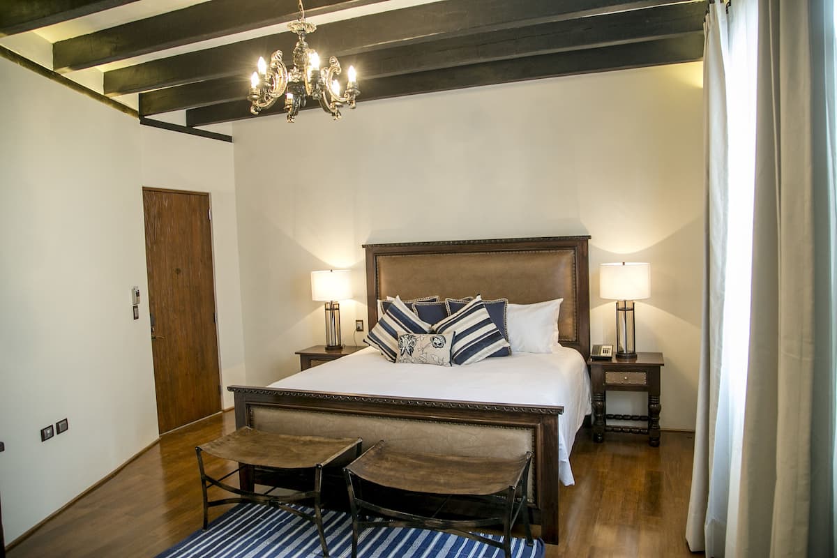 Quarto de hotel com uma cama grande, duas mesas de cabeceira com luminárias, um candelabro, uma porta de madeira e duas cadeiras.