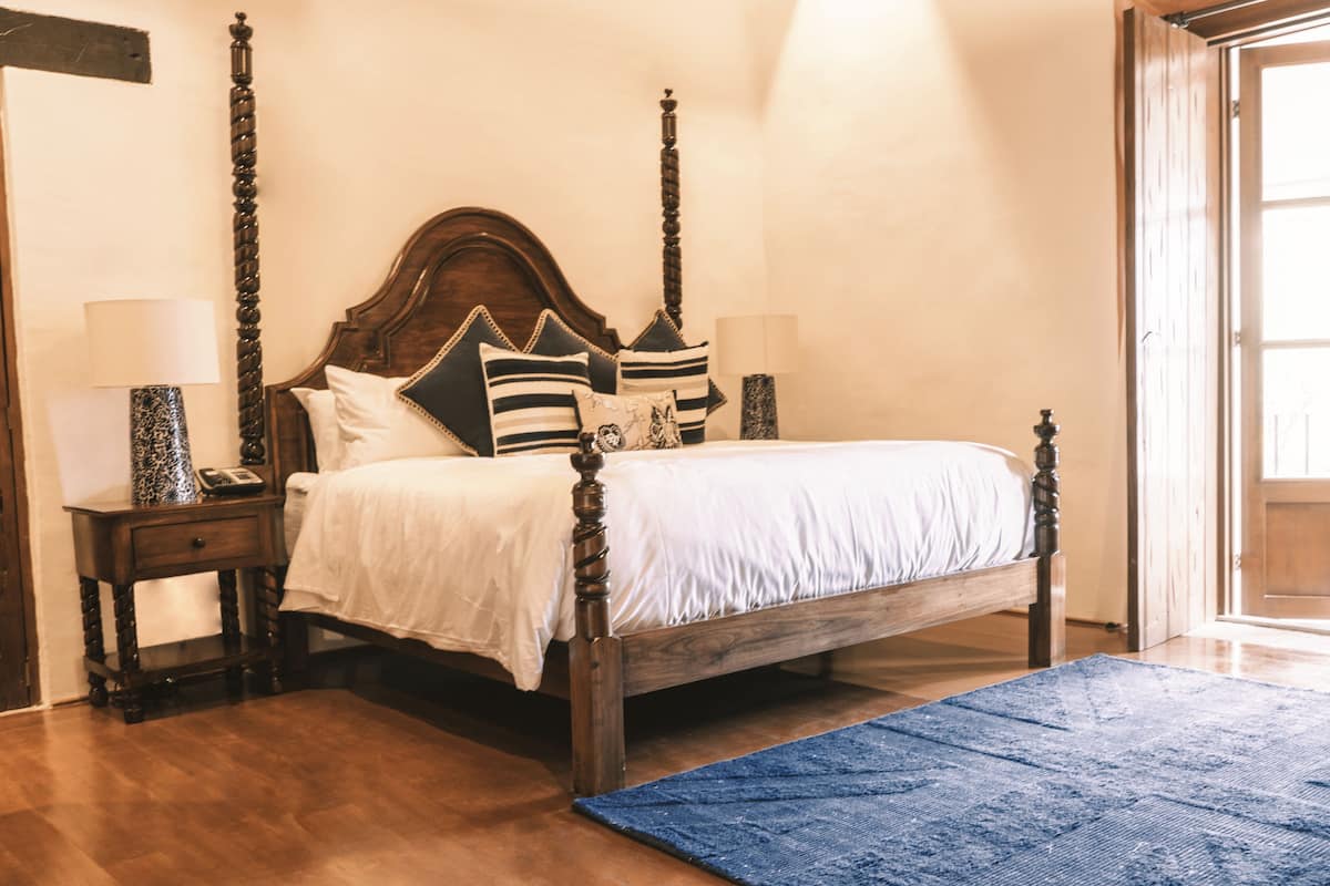 Quarto com cama de dossel de madeira, roupa de cama branca, almofadas listradas, duas mesas de cabeceira com luminárias e um tapete azul.