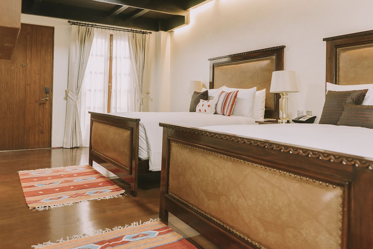 Quarto de hotel com duas camas de madeira, uma mesa de cabeceira, um abajur e um tapete estampado.