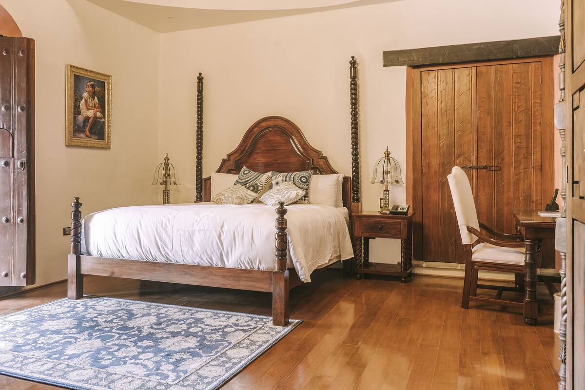 Quarto com uma cama de dossel de madeira, mesa de cabeceira com abajur, uma cadeira e guarda-roupa de madeira.