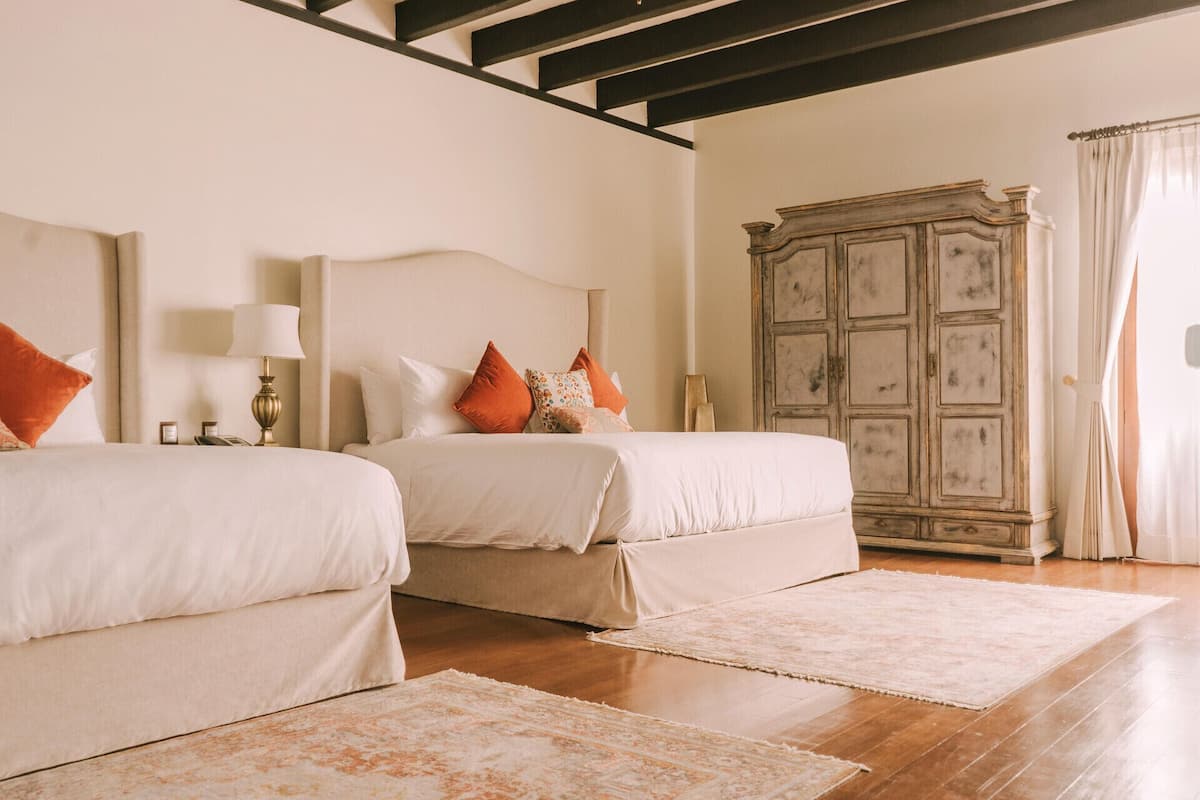 Quarto de hotel com duas camas, um guarda-roupa de madeira e uma janela grande com cortinas.