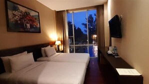 Quarto de hotel com duas camas, uma escrivaninha com televisão de tela plana, um quadro na parede e vista para uma piscina à noite.