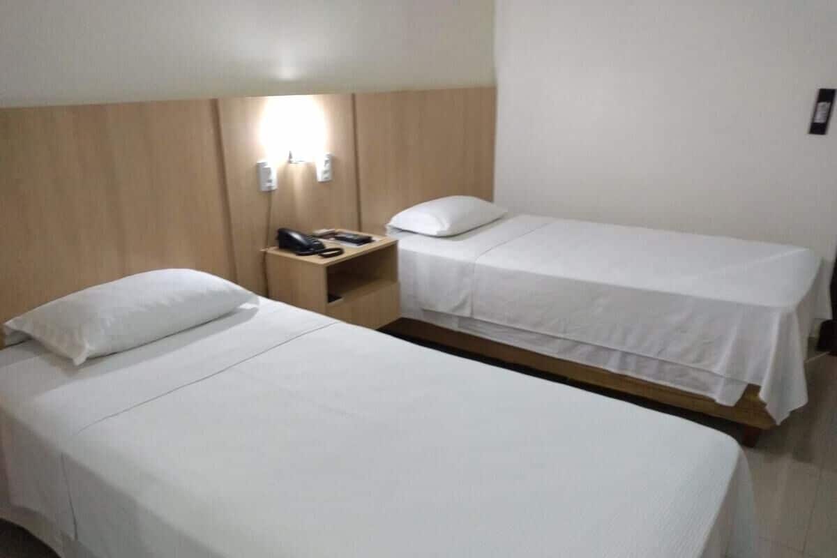 Quarto de hotel com duas camas de solteiro, uma mesa de cabeceira com luminária e uma tomada na parede.