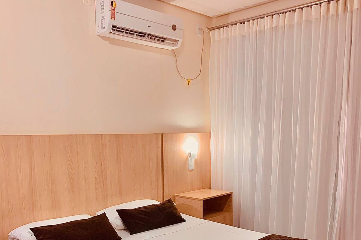 Quarto de hotel com cama, mesa de cabeceira, ar condicionado fixado na parede e janela com cortinas.