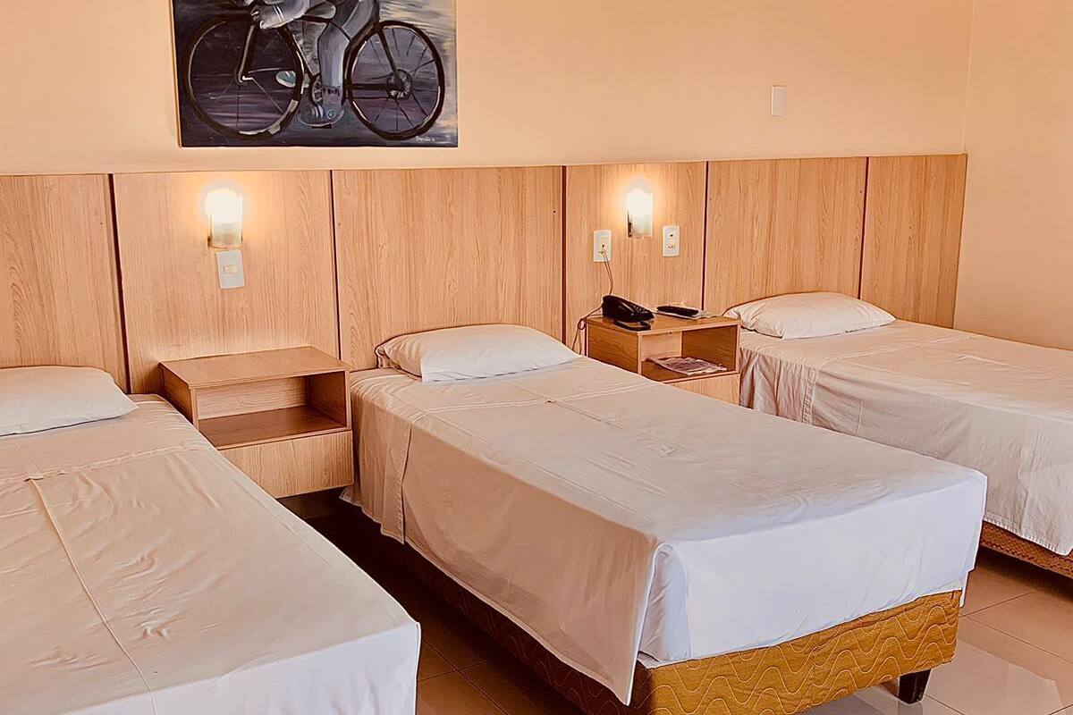 Quarto de hotel com três camas de solteiro, cabeceiras de madeira, um quadro na parede retratando um ciclista e uma mesa de cabeceira com abajur.