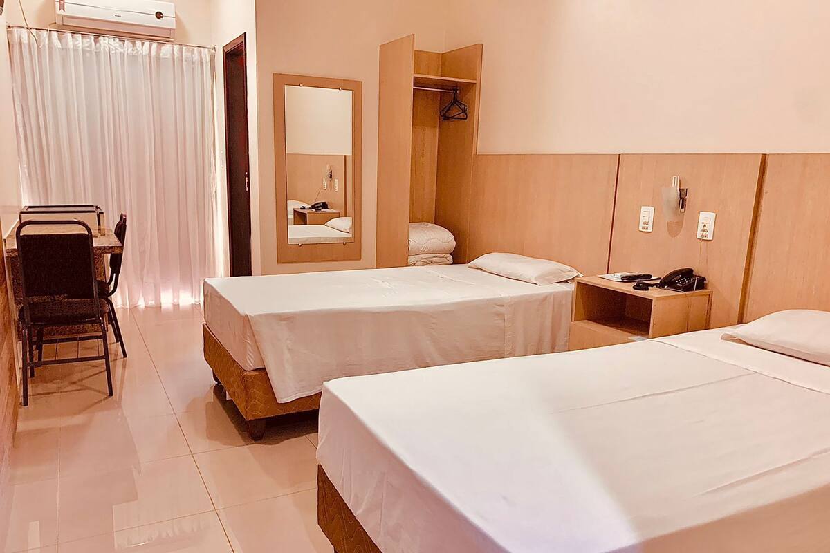 Quarto de hotel com duas camas, uma escrivaninha, uma cadeira, um guarda-roupa e um espelho.
