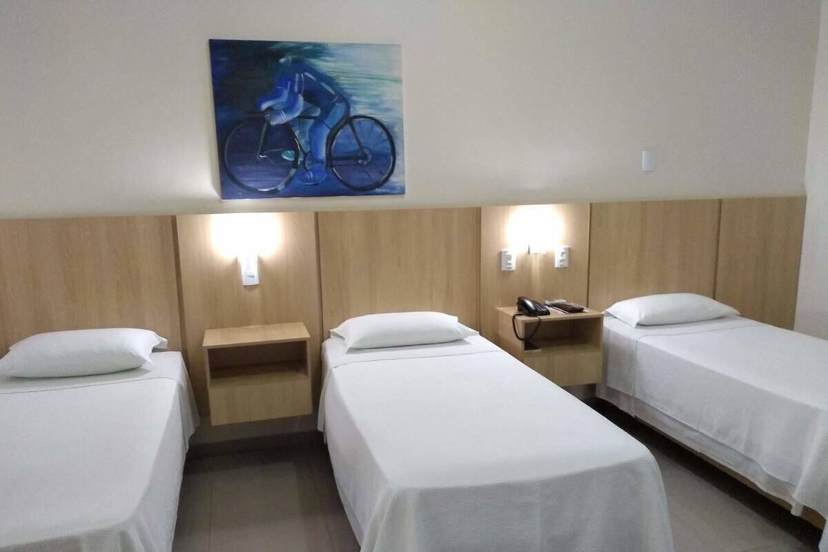 Quarto de hotel com três camas de solteiro, cabeceiras de madeira, mesas de cabeceira e um quadro pendurado na parede retratando um ciclista.