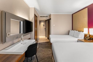 Quarto de hotel com cama, escrivaninha, cadeira, televisão, espelho e mesa de cabeceira com abajur.