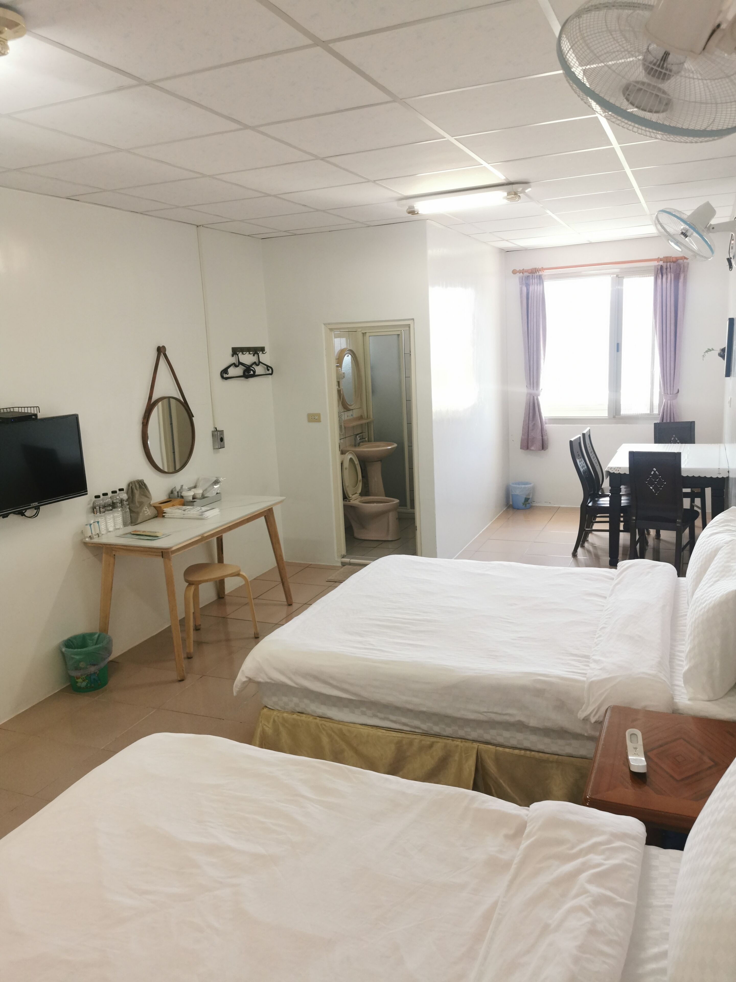 Quarto de hotel com duas camas, uma escrivaninha, uma cadeira, televisão, espelho e janela com cortinas.