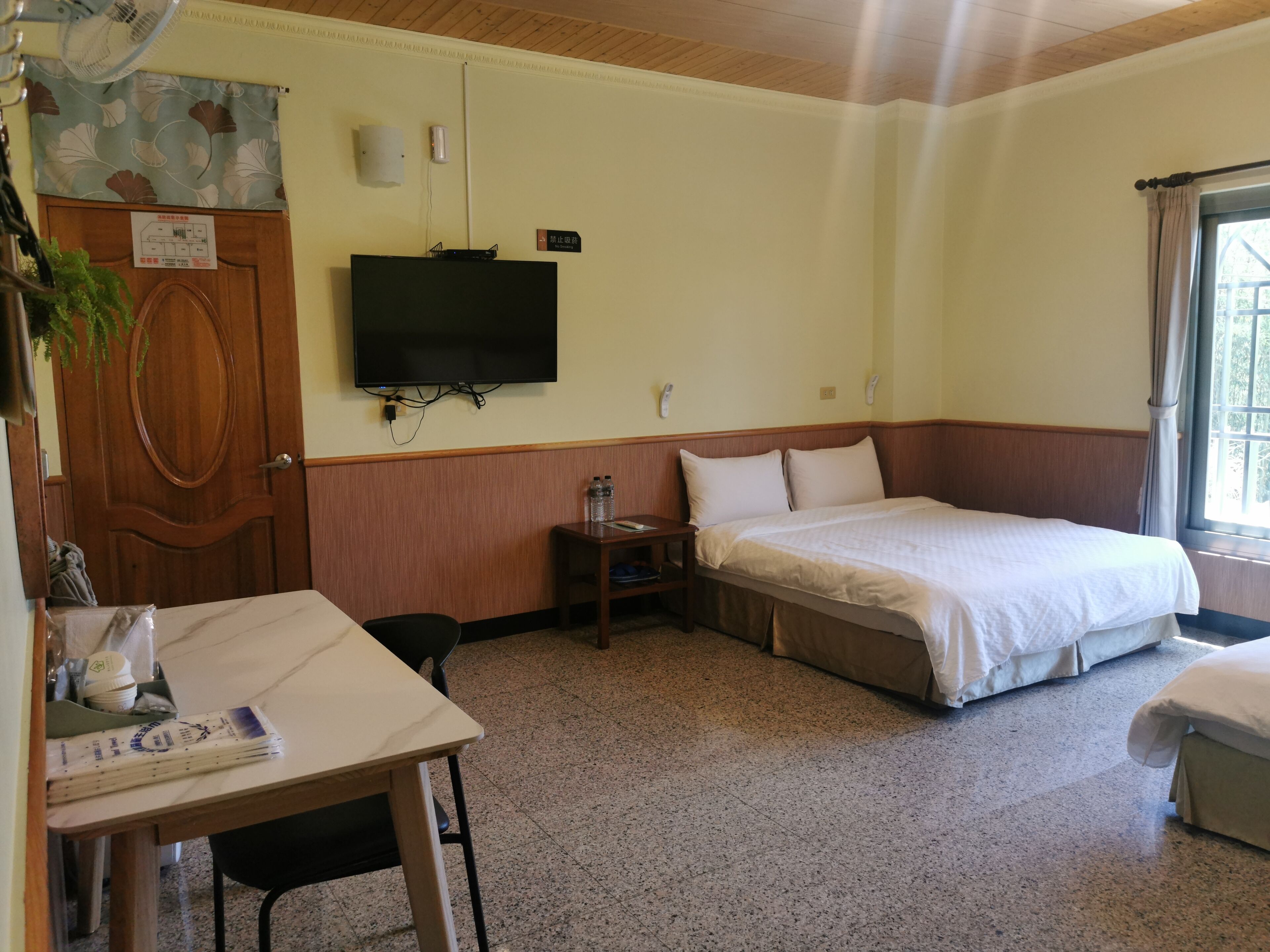 Quarto de hotel com cama, escrivaninha, cadeira, televisão e janela.