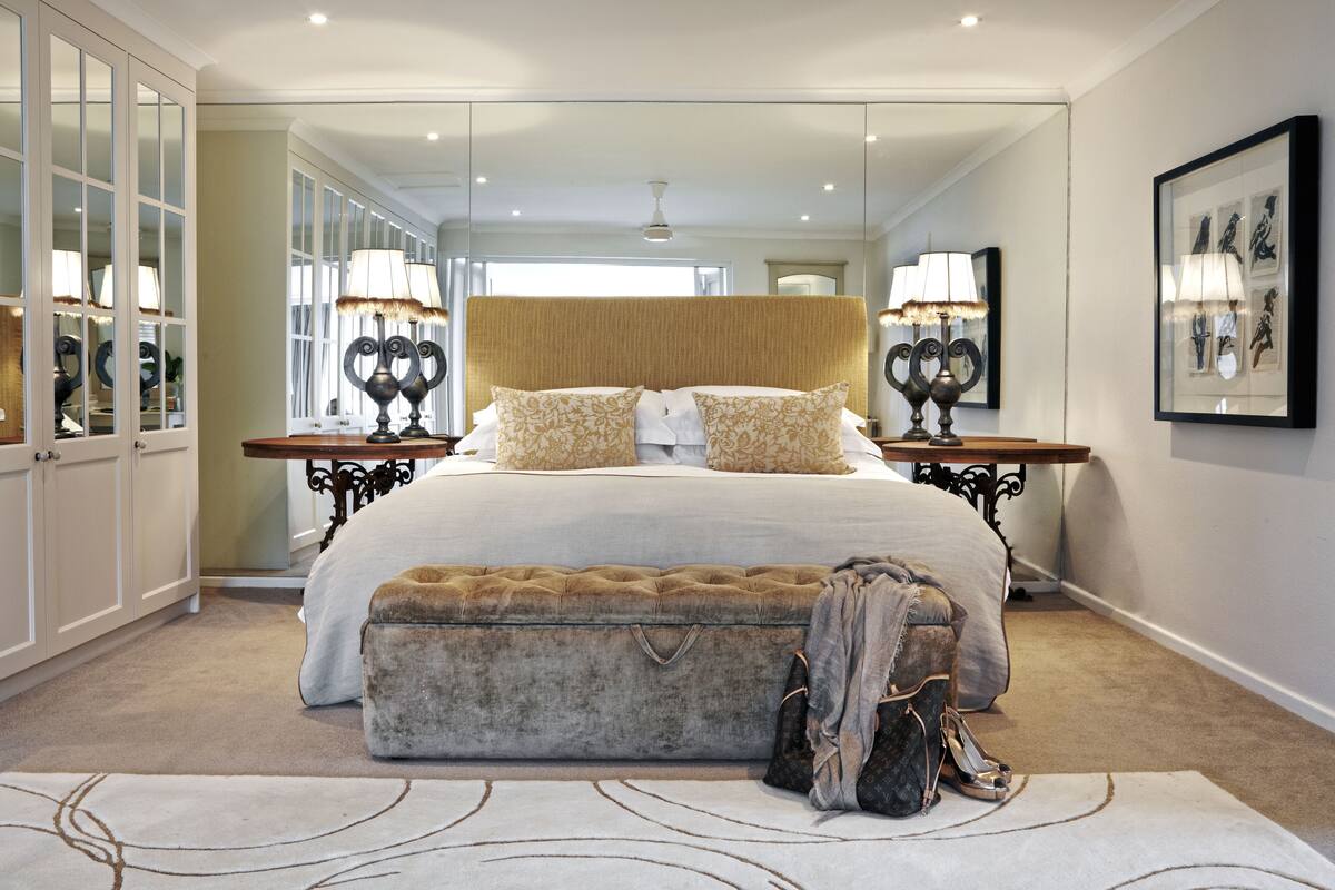 Um quarto espaçoso com uma cama grande, um banquinho estofado e um espelho refletindo o ambiente.