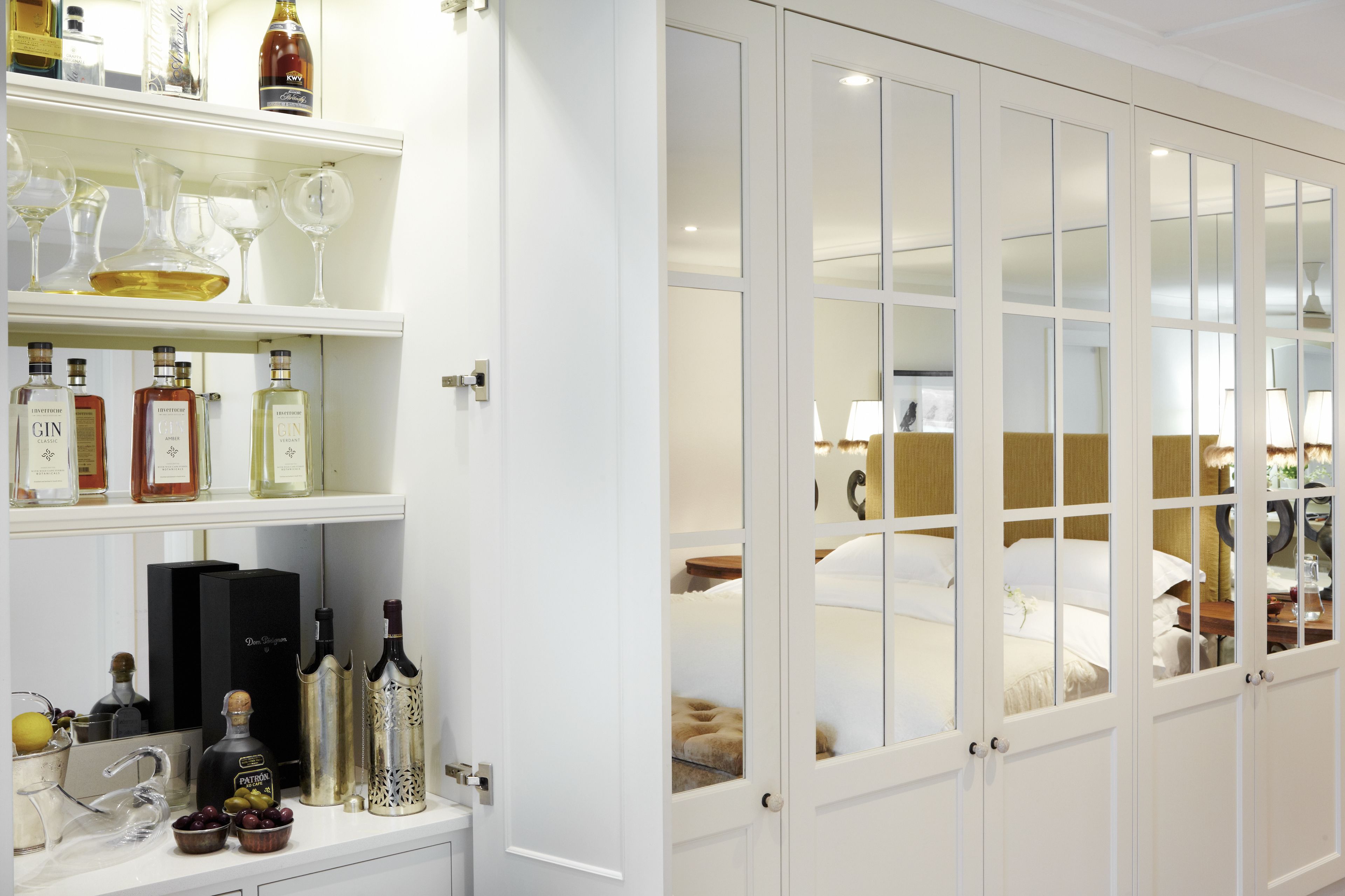 Quarto de hotel com minibar, cama com lençóis brancos e um espelho grande refletindo o ambiente.