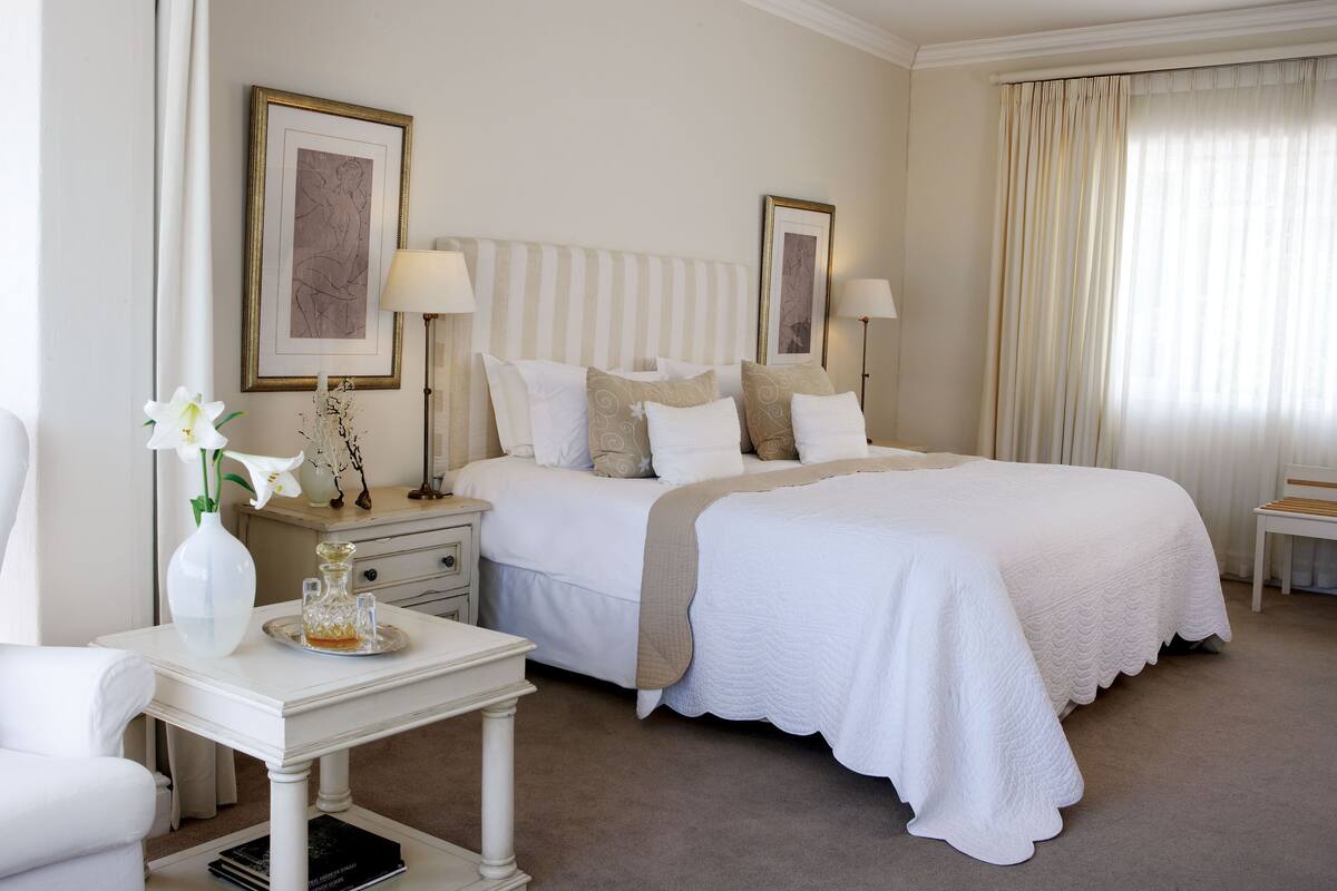 Quarto de hotel com uma cama grande, mesa de cabeceira com abajur, uma cadeira e um vaso com flores.