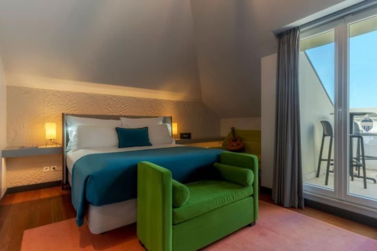 Quarto de hotel com cama, uma cadeira verde, uma janela grande e varanda.