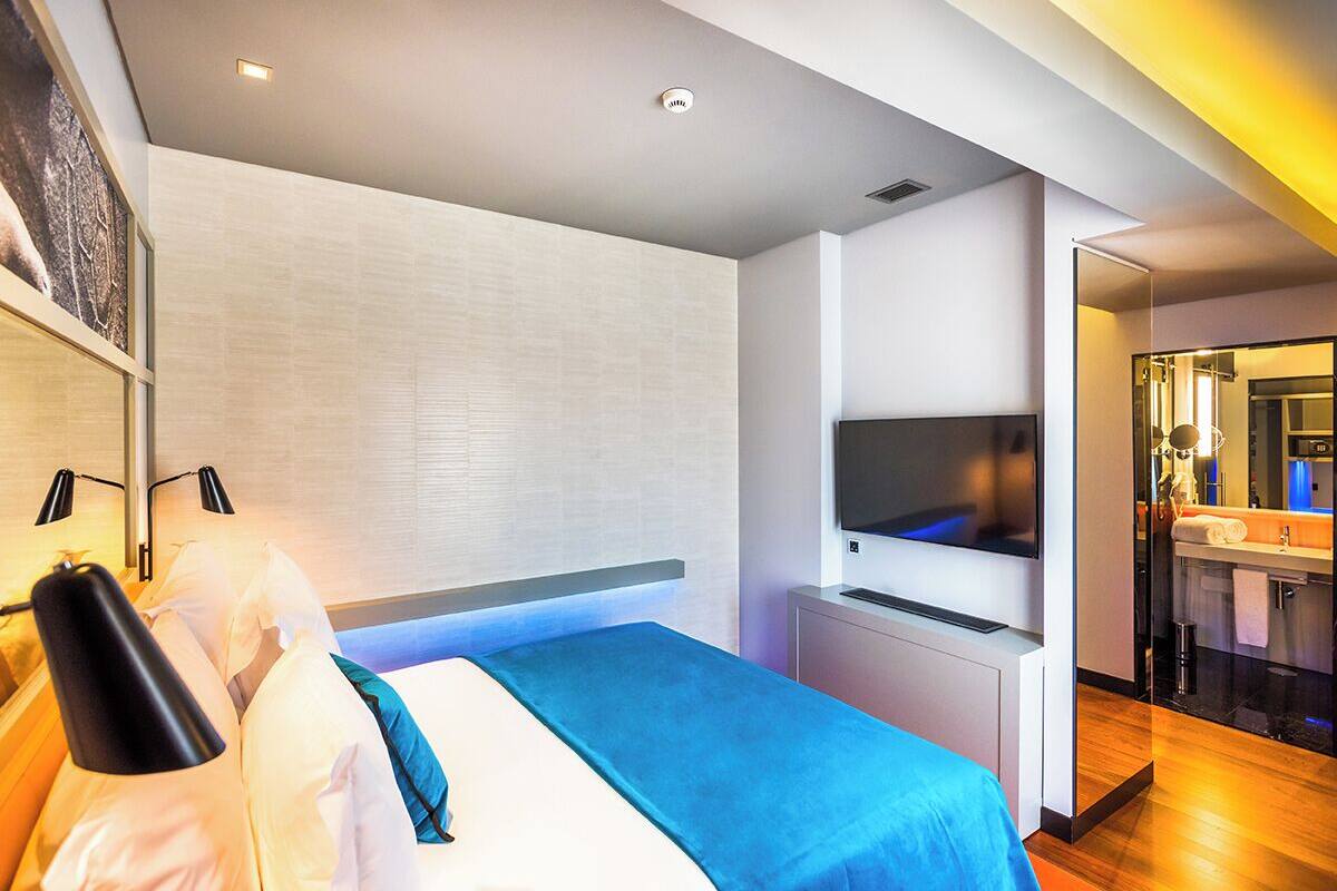 Quarto de hotel moderno com uma cama, uma televisão de tela plana, uma escrivaninha e um banheiro visível por uma porta de vidro.