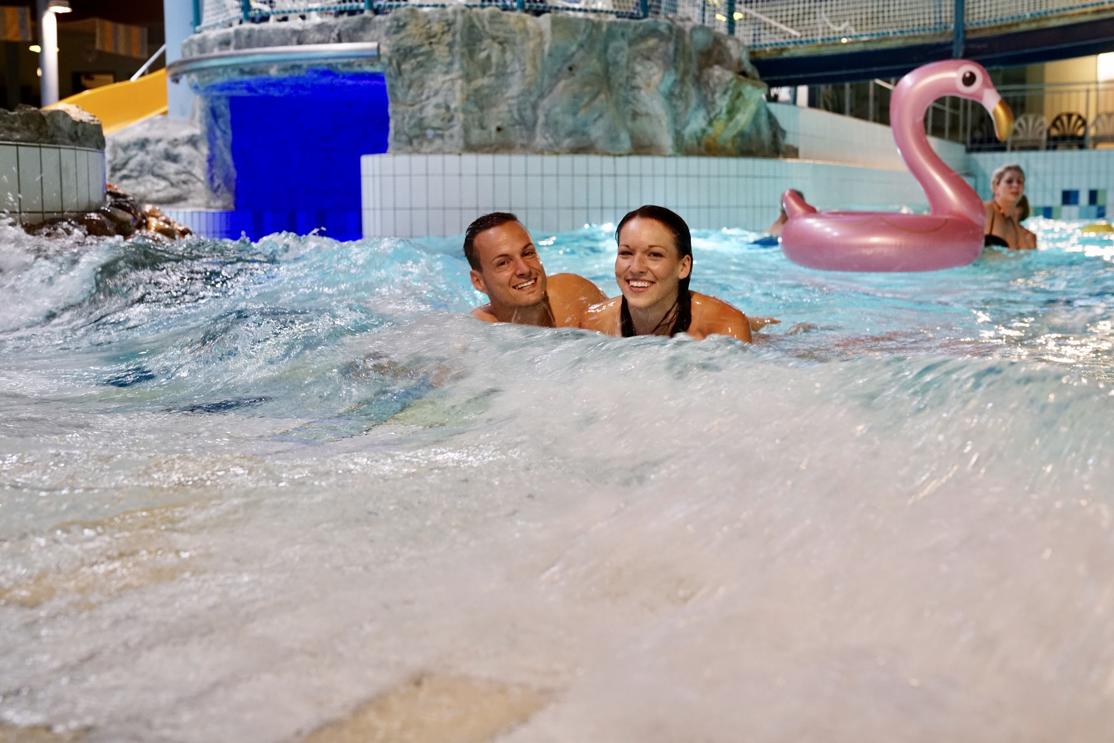 Um casal em banheira de hidromassagem com um flutuador de cisne rosa, cercado por elementos aquáticos e uma parede revestida de azulejos.