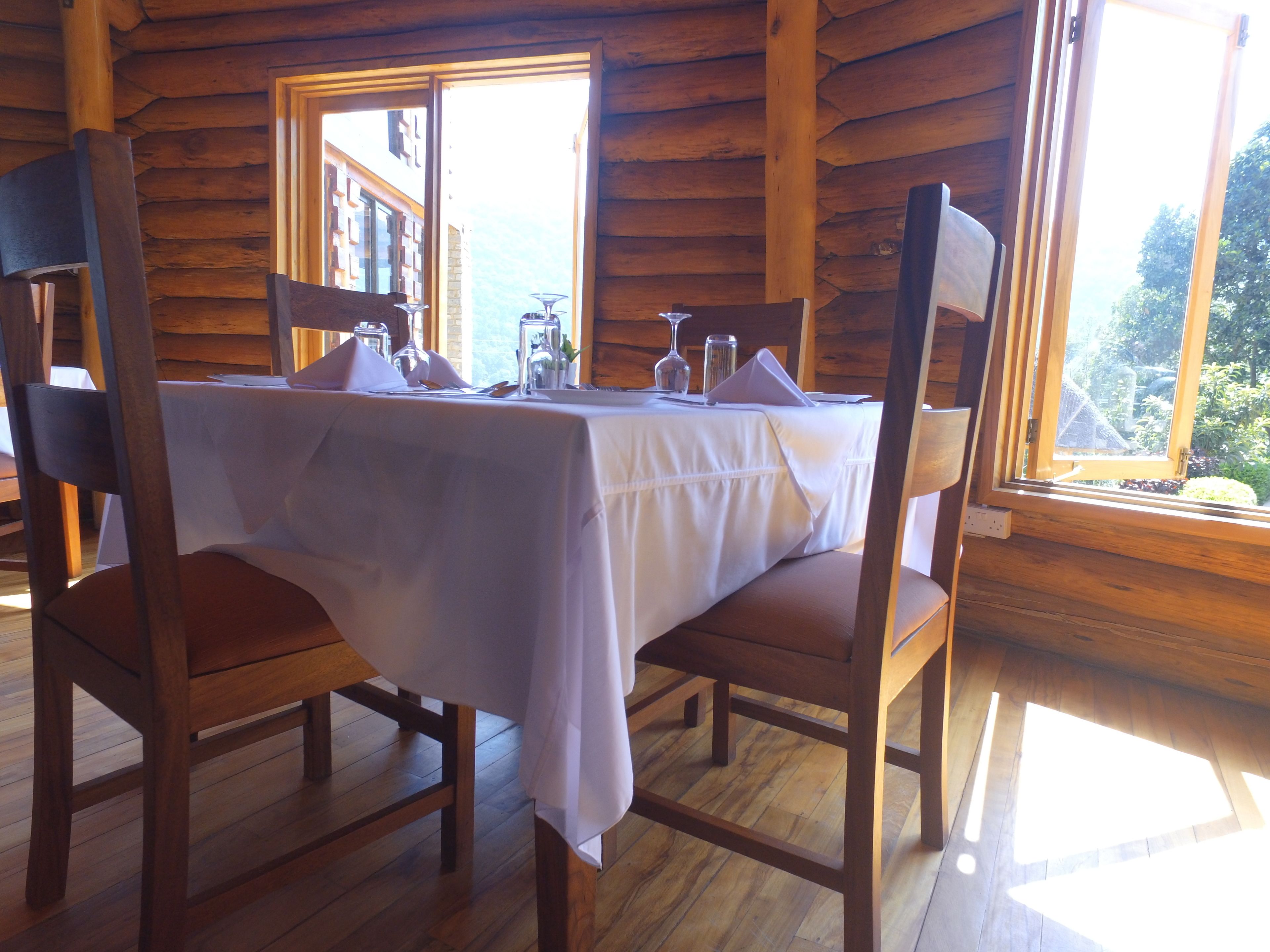 Interior de uma cabana de madeira, com uma mesa de jantar posta para dois, cadeiras de madeira e uma janela com vista para o verde.