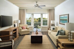 Quarto de hotel com televisão de tela grande, dois sofás, uma mesa de centro, ventilador de teto e varanda com cortinas.
