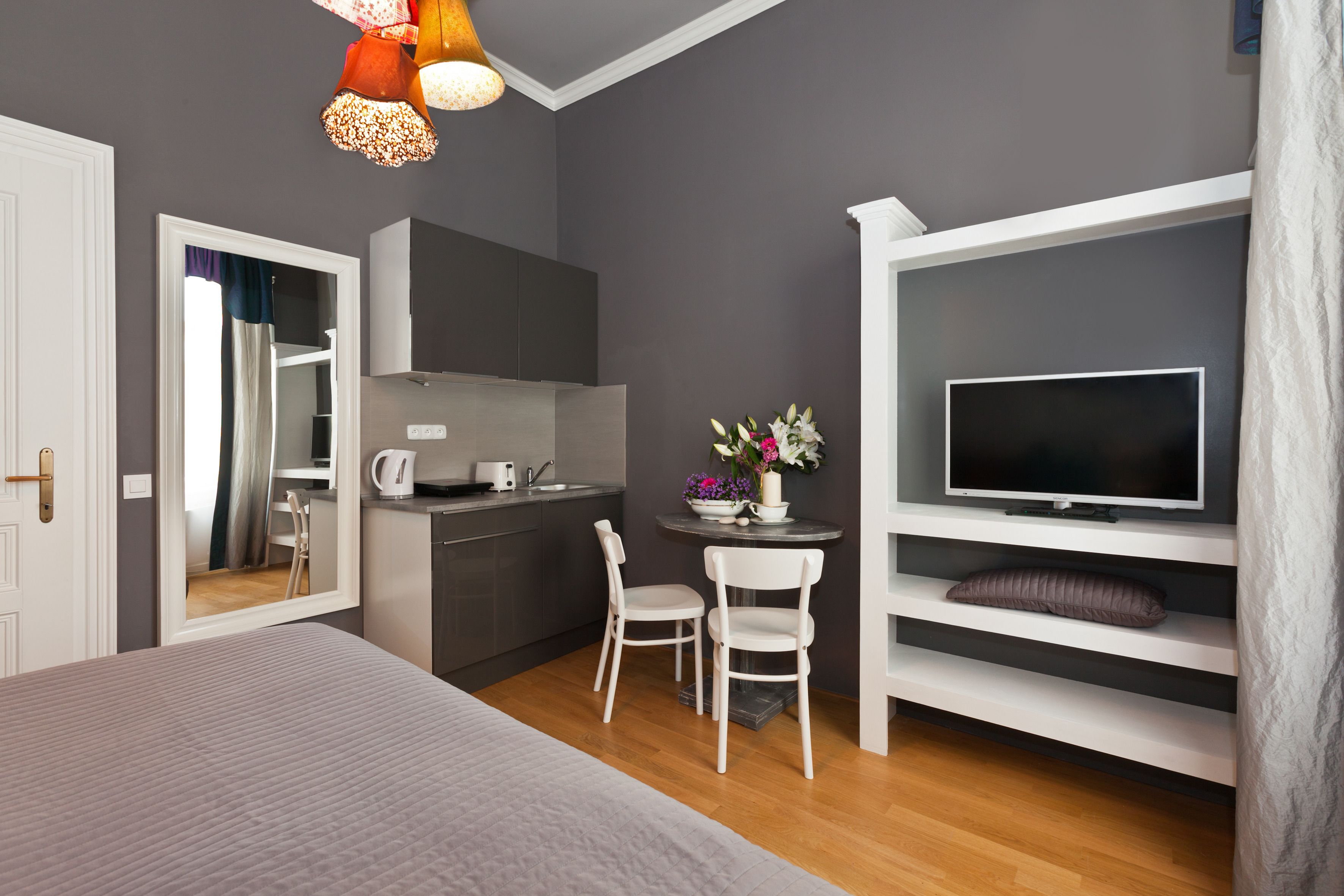 Quarto de hotel moderno com cama, televisão sobre uma estante branca, pequena área de cozinha e espelho.