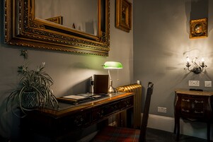 Quarto com escrivaninha de madeira, luminária de mesa verde, radiador, cadeira xadrez e um quadro pendurado na parede.