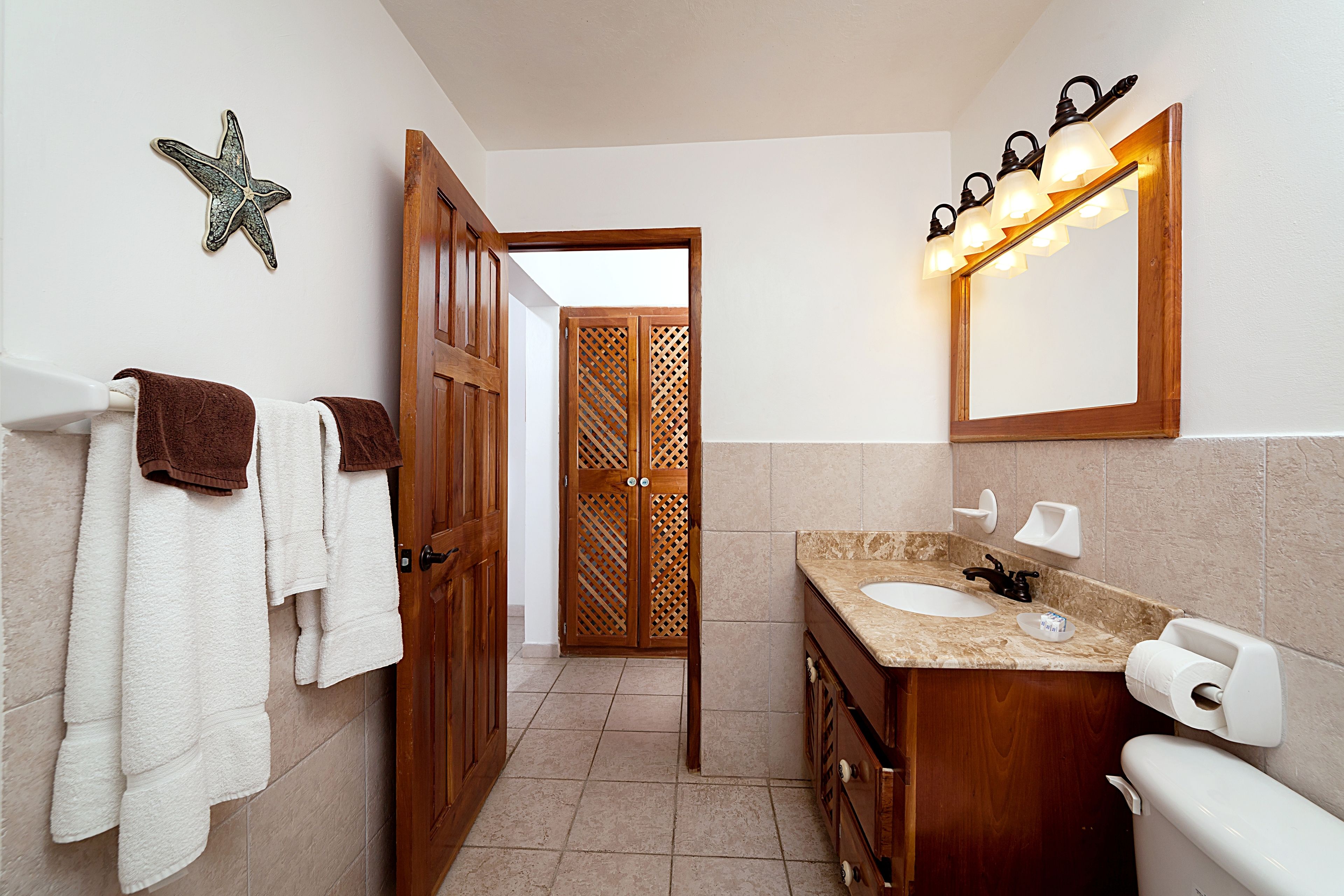 Casa de banho com toucador de madeira, lavatório, sanita e espelho com luzes. Toalhas penduradas na parede e um adorno em forma de estrela.